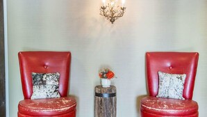 Duas poltronas de couro vermelho com almofadas estampadas, uma mesinha de madeira com um vaso de flores e um candelabro fixado na parede.
