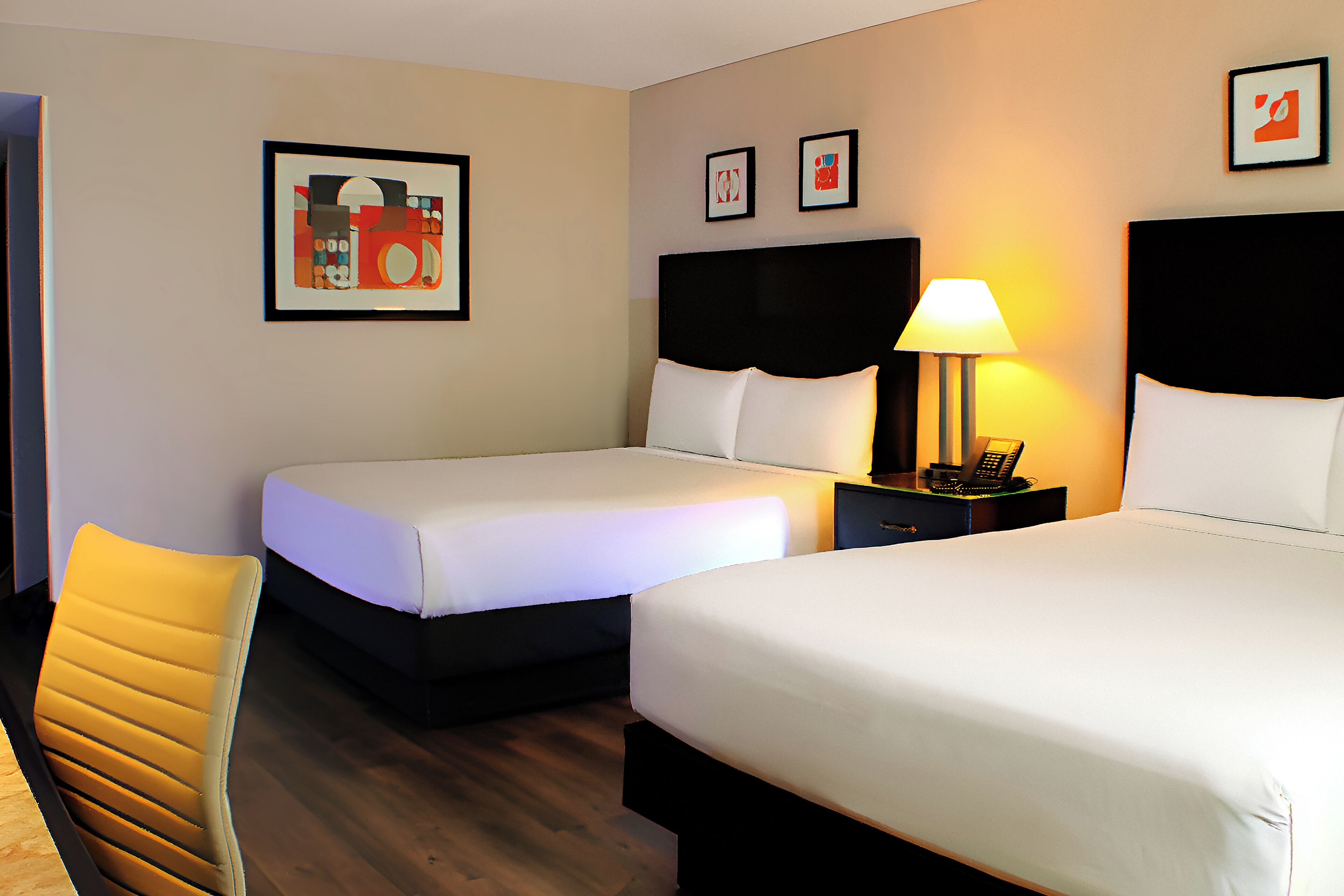 Quarto de hotel com duas camas, uma cadeira e uma mesa de cabeceira com abajur.