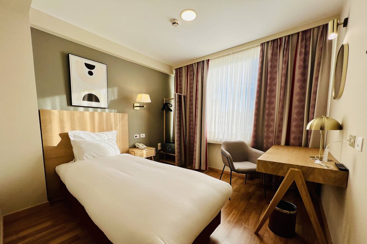 Quarto de hotel com cama, escrivaninha, cadeira, abajur, telefone e janela com cortinas.