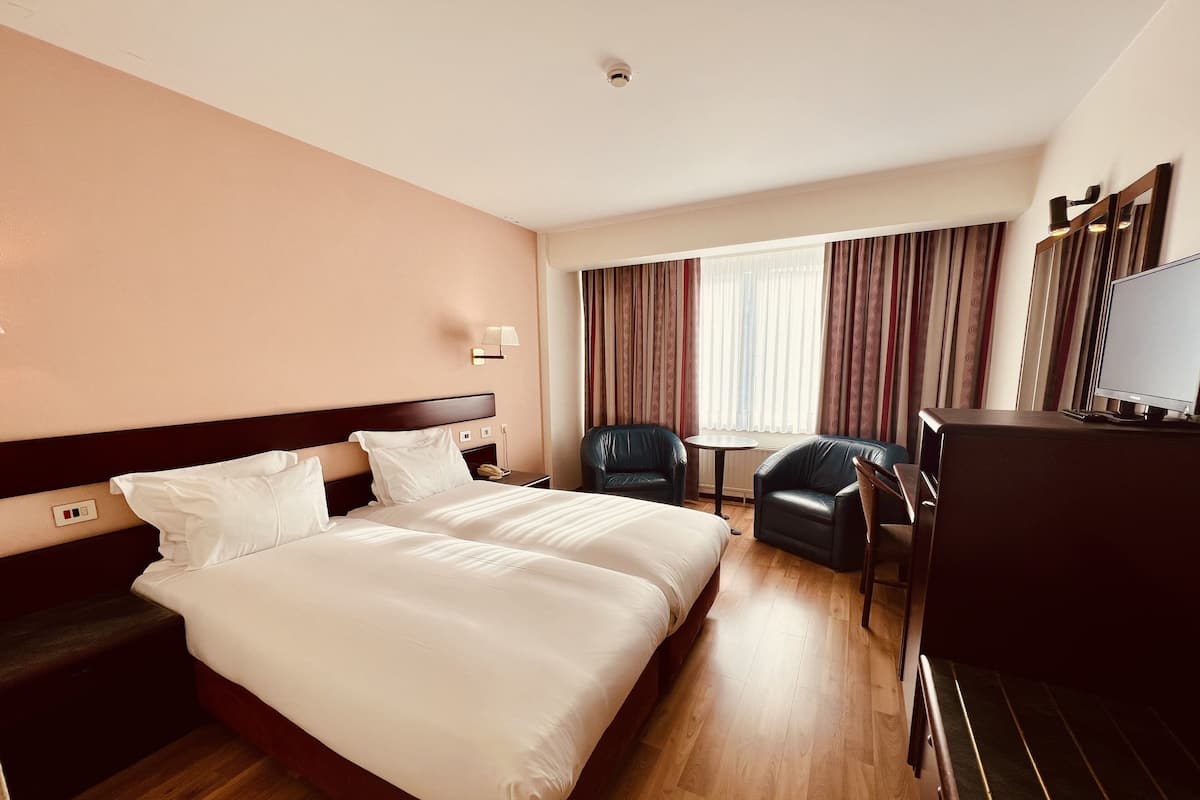 Quarto de hotel com uma cama grande, duas cadeiras, uma escrivaninha e uma televisão.