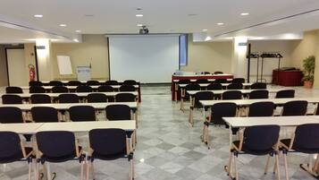 Uma sala de conferências com fileiras de mesas e cadeiras, uma tela de projetor, um púlpito e um extintor de incêndio.