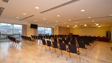 Sala de conferências com fileiras de cadeiras, uma janela grande com vista para edifícios e uma TV fixada na parede.