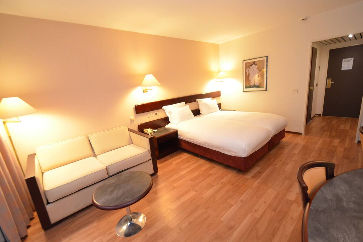 Quarto de hotel com cama, sofá, uma mesinha e duas luminárias de parede.
