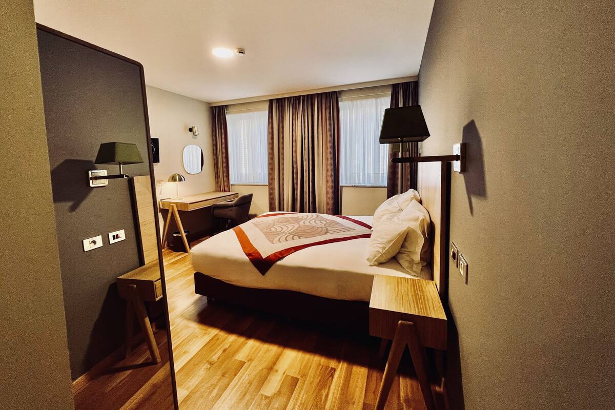 Quarto de hotel com cama, escrivaninha, cadeira e janela com cortinas.