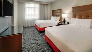 Quarto de hotel com duas camas, televisão de tela plana, mesa de cabeceira com abajur e uma janela grande com cortinas.