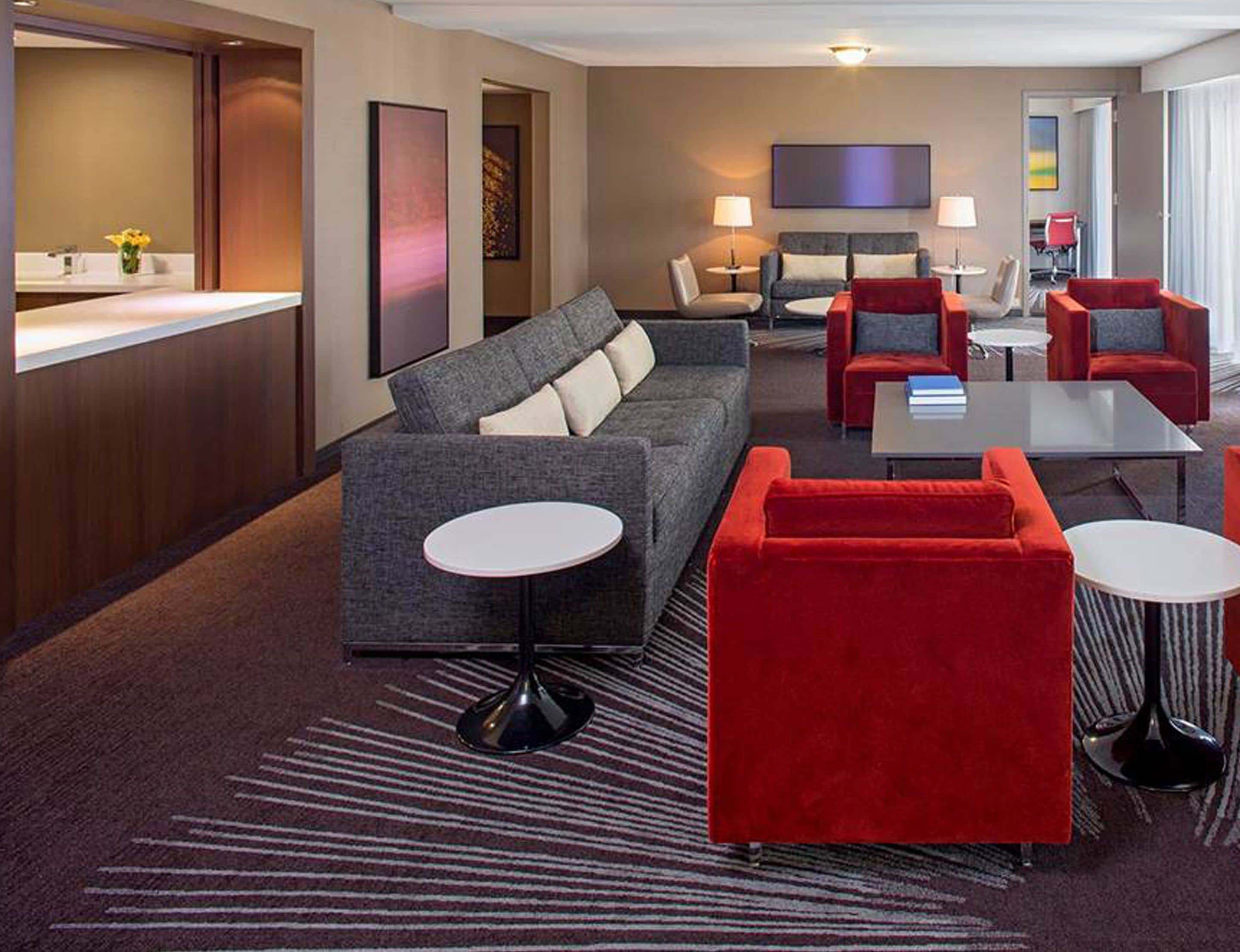 Quarto de hotel moderno com sofá cinzento, poltronas vermelhas, uma mesa pequena e televisão na parede.