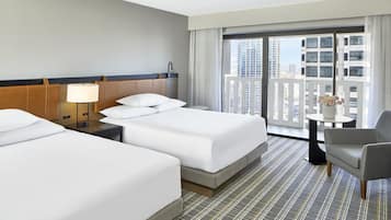 Quarto de hotel com duas camas, uma mesa de cabeceira com abajur, uma pequena mesa com um vaso e uma cadeira. O quarto possui uma janela grande com vista para edifícios.