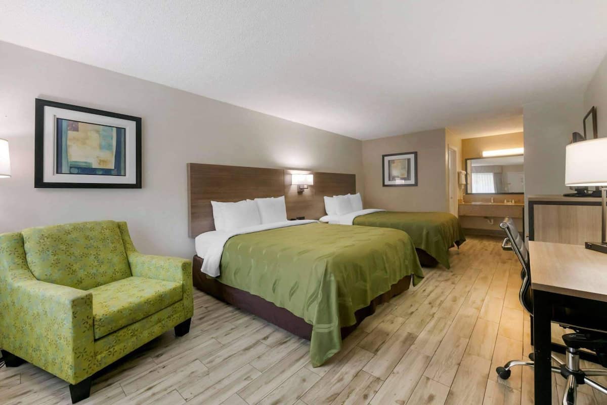 Quarto de hotel com duas camas, uma poltrona verde, uma escrivaninha com cadeira e um espelho.