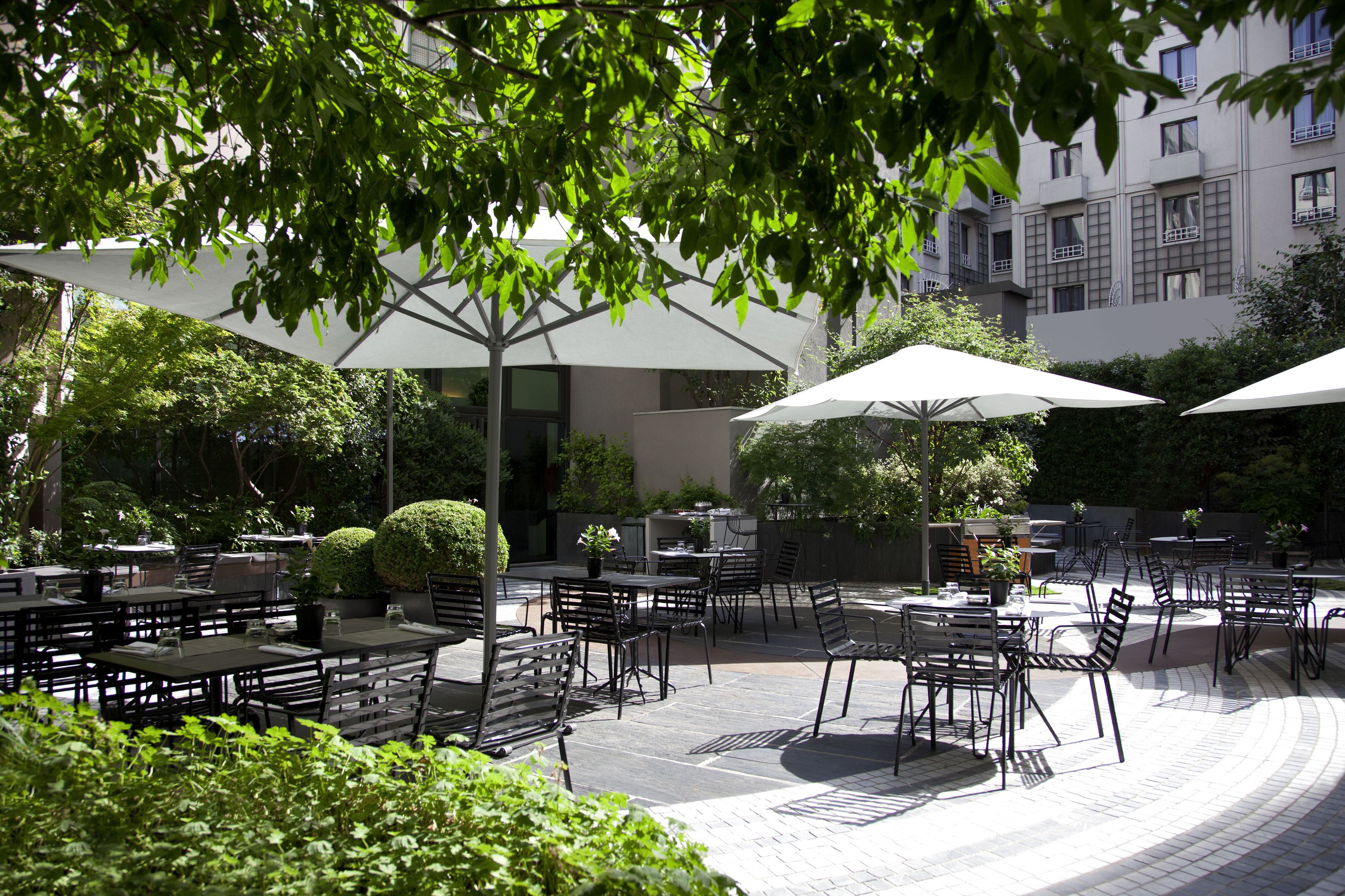 Um terraço ao ar livre com mesas, cadeiras, guarda-sóis e vegetação.