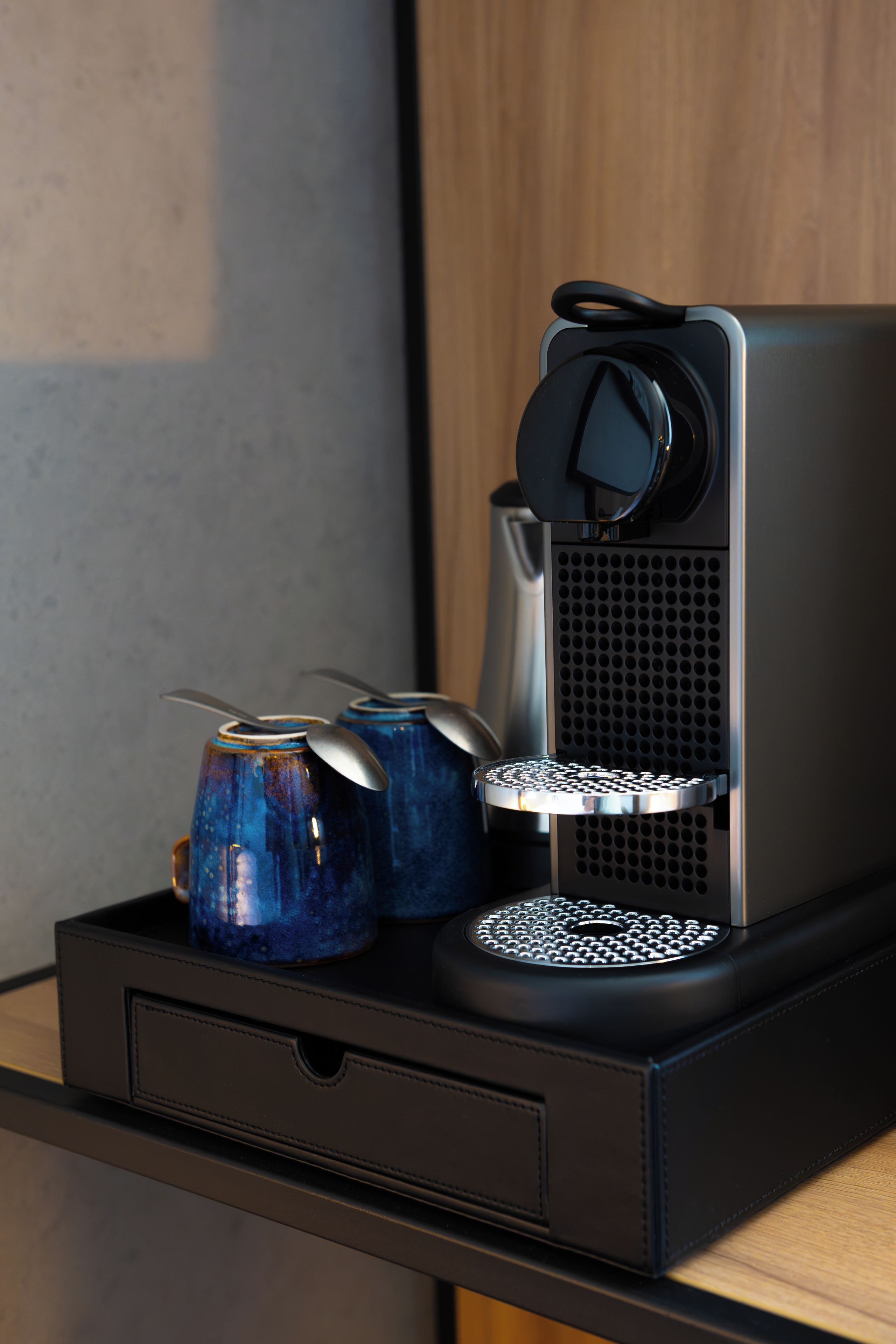 Máquina de café com bandeja, duas chávenas azuis e uma colher sobre uma superfície preta.