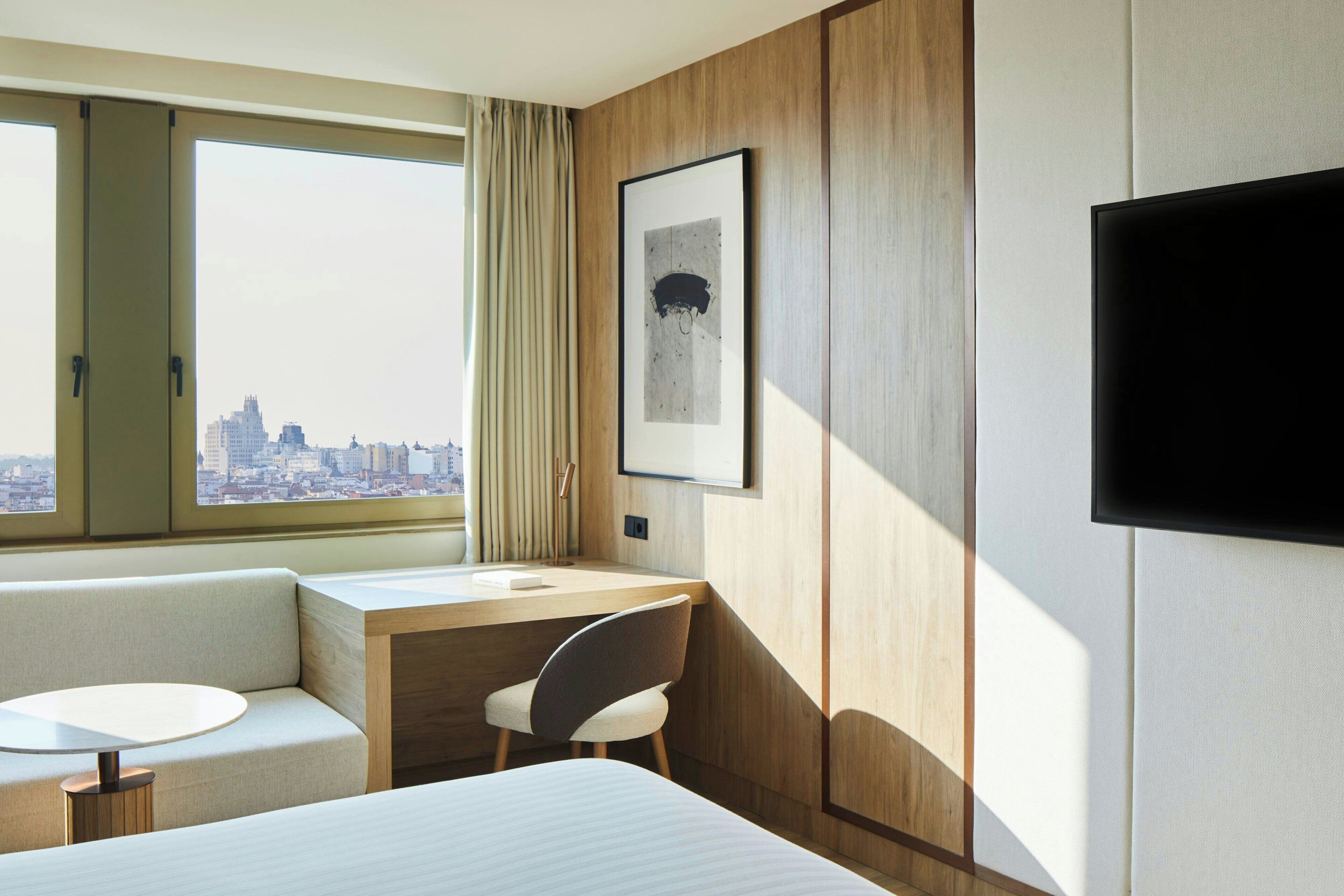 Quarto de hotel com cama, escrivaninha, cadeira, mesa pequena, televisão e janela com vista para a cidade.