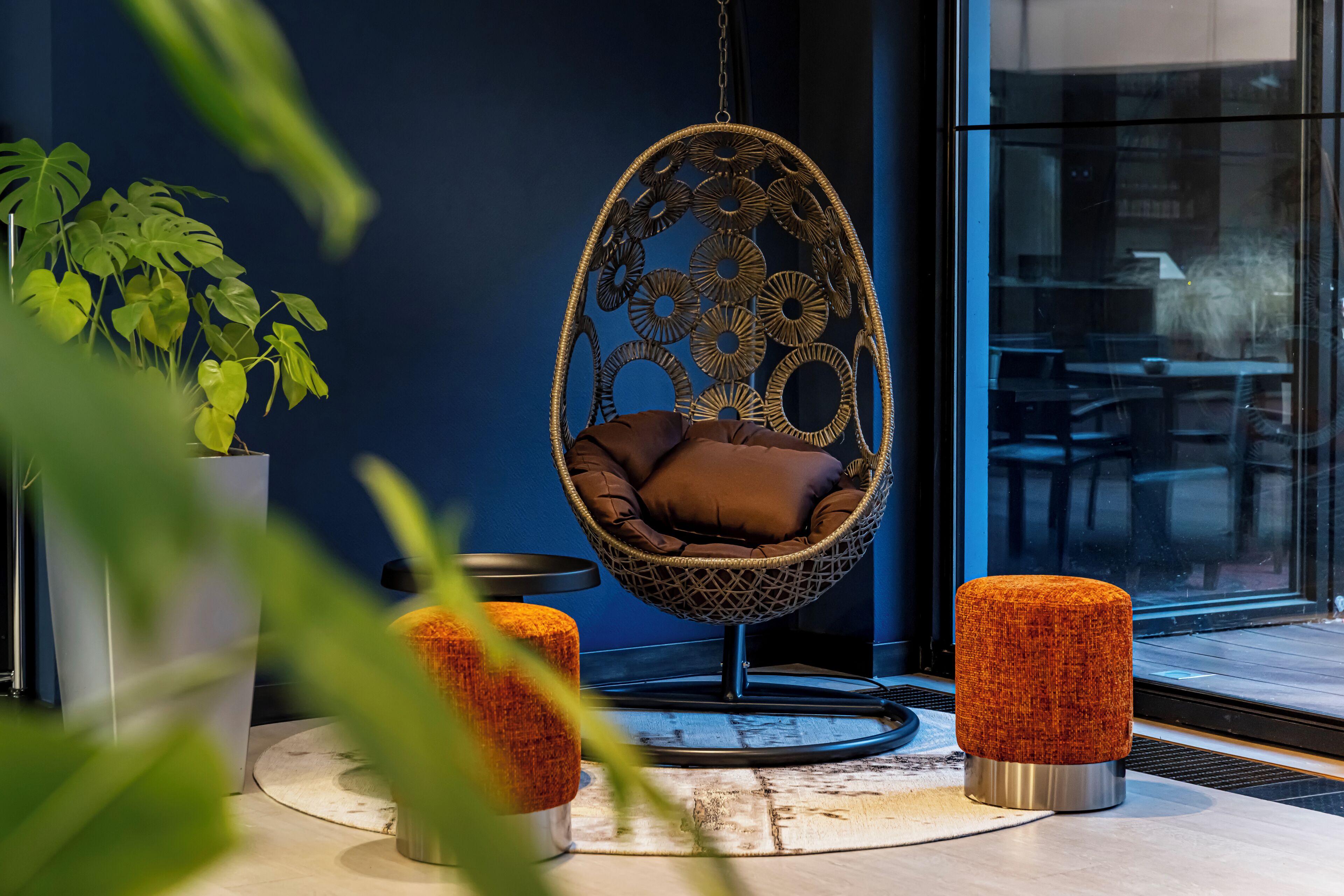 Uma poltrona suspensa com almofada, dois pufes e um vaso com planta em um espaço interior moderno.