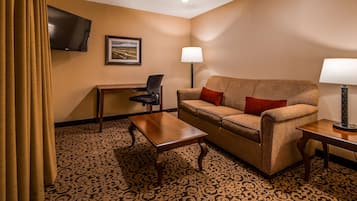 Quarto de hotel com sofá, escrivaninha, cadeira, televisão, luminária e piso coberto de carpete.