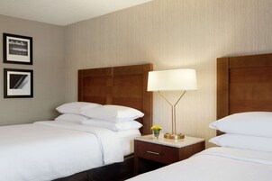 Quarto de hotel com duas camas, uma mesa de cabeceira com abajur e quadros nas paredes.