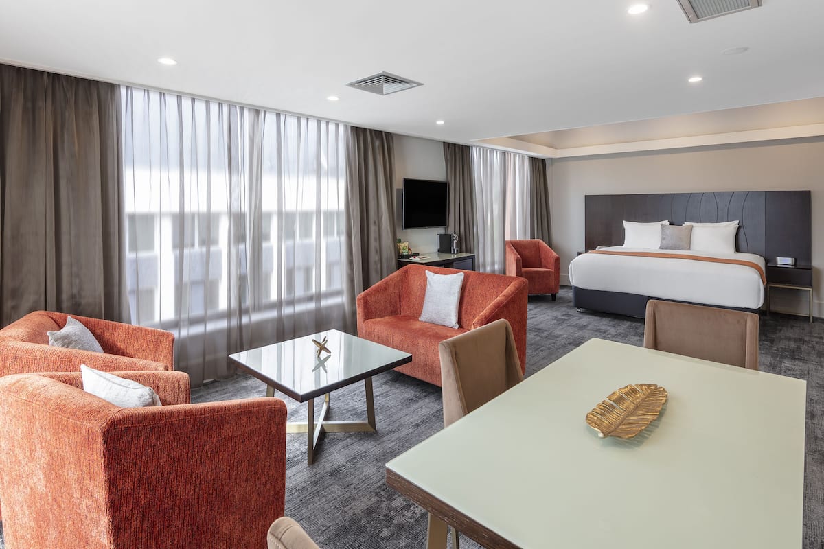 Quarto de hotel com cama, duas poltronas laranja, uma mesa pequena, televisão e uma janela grande com cortinas.