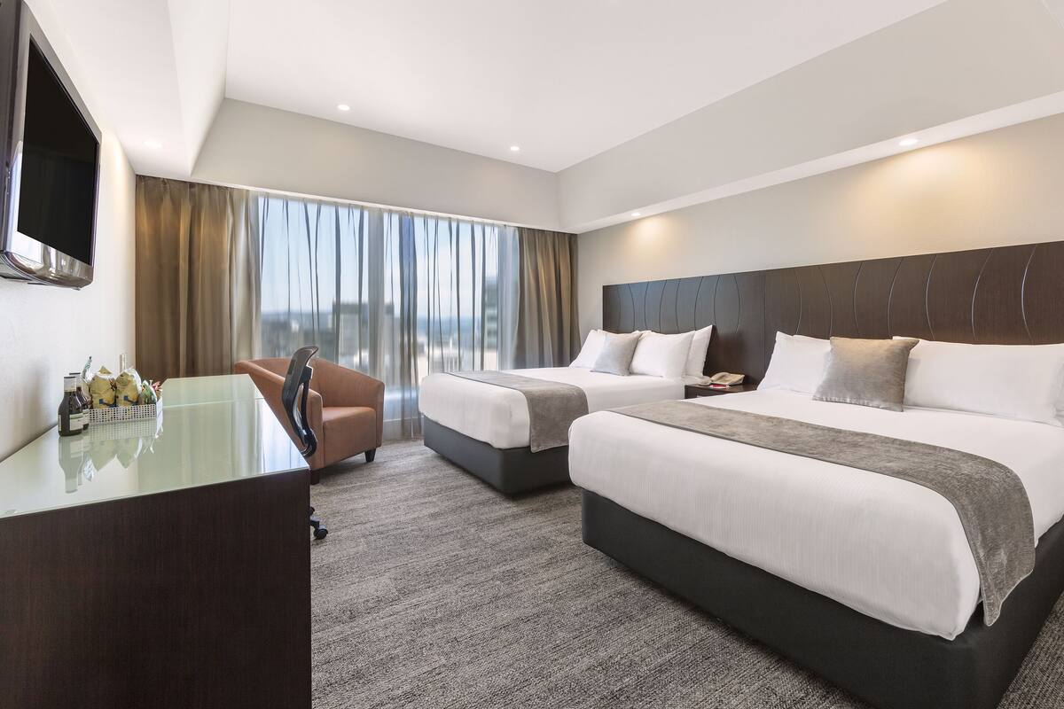 Quarto de hotel com uma cama grande, uma cama menor, uma escrivaninha com televisão, uma cadeira e vista da cidade por grandes janelas.
