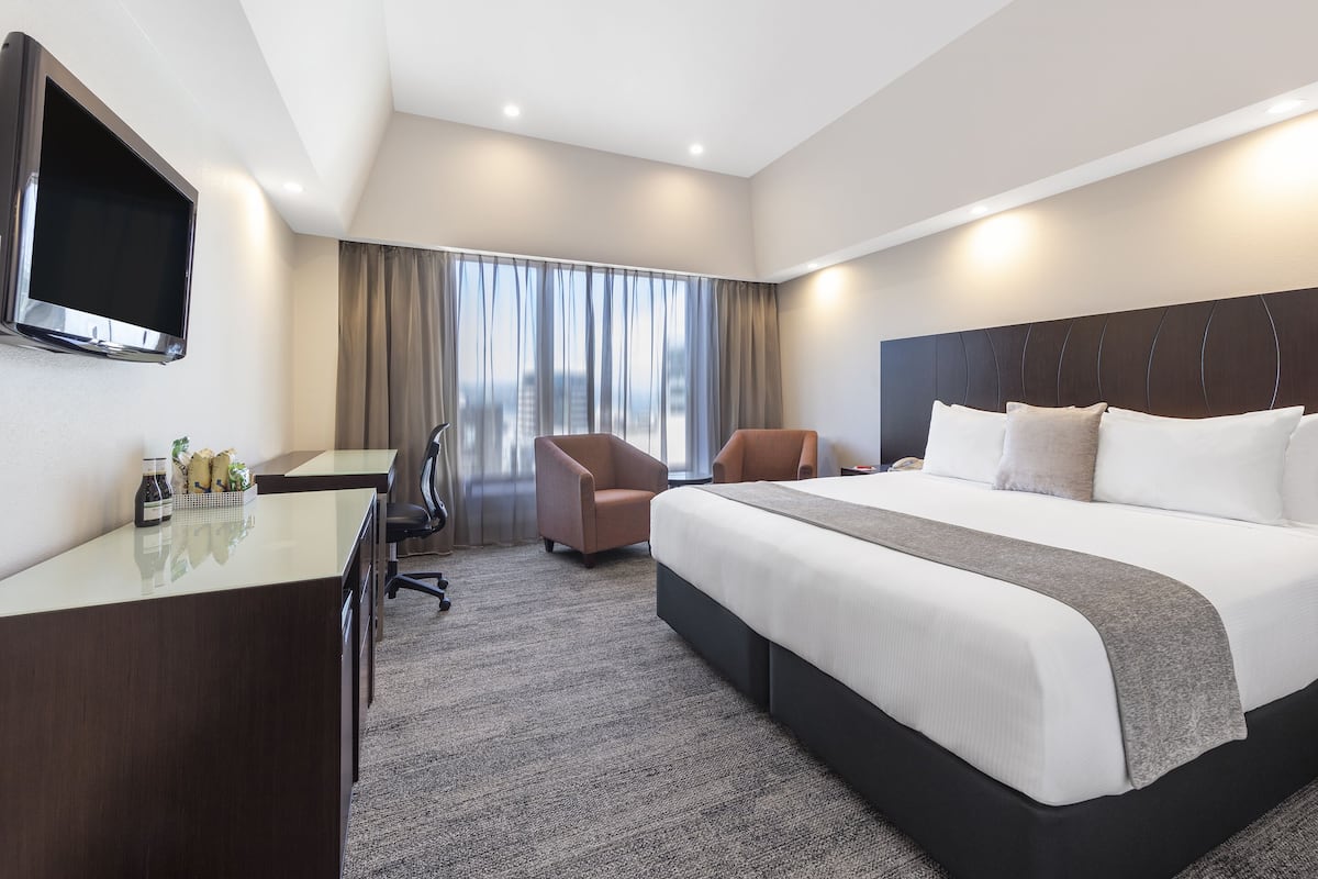 Quarto de hotel com uma cama grande, uma escrivaninha, uma cadeira, uma TV fixada na parede e vista para a cidade pela janela.