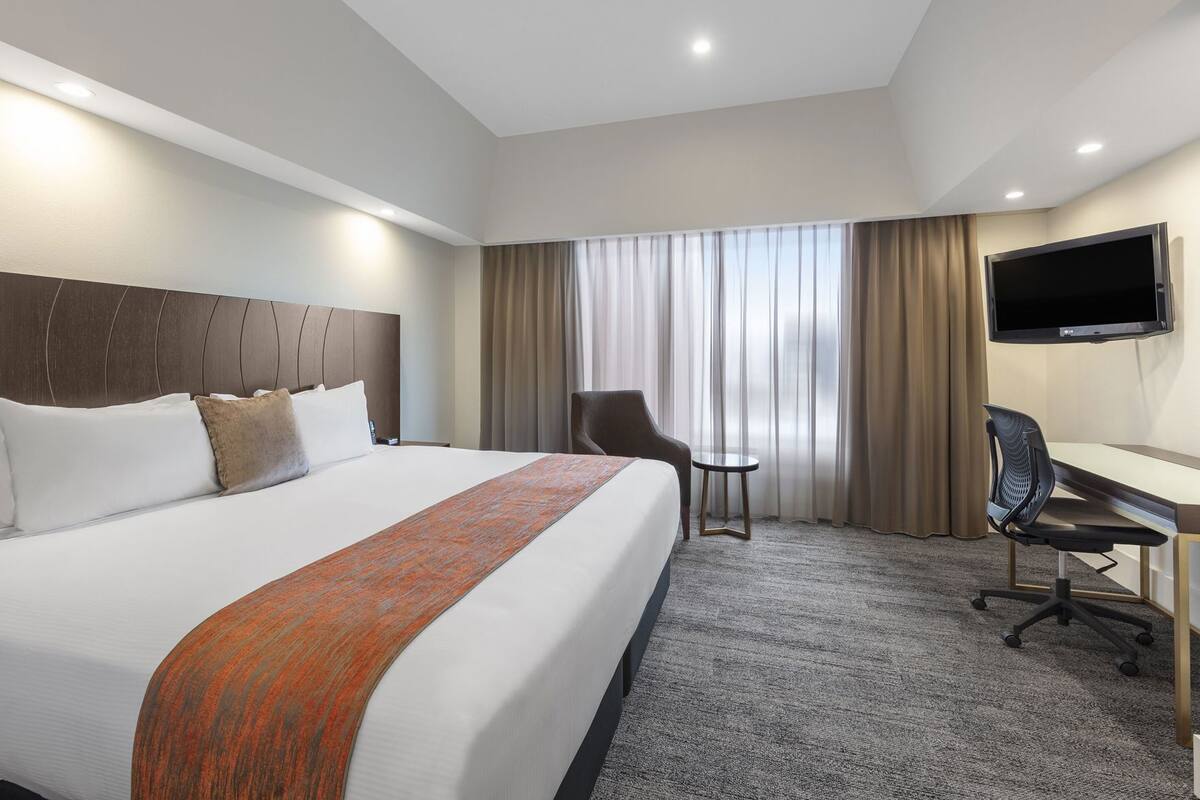 Quarto de hotel com uma cama grande, uma mesa com cadeira, televisão e janela com cortinas.