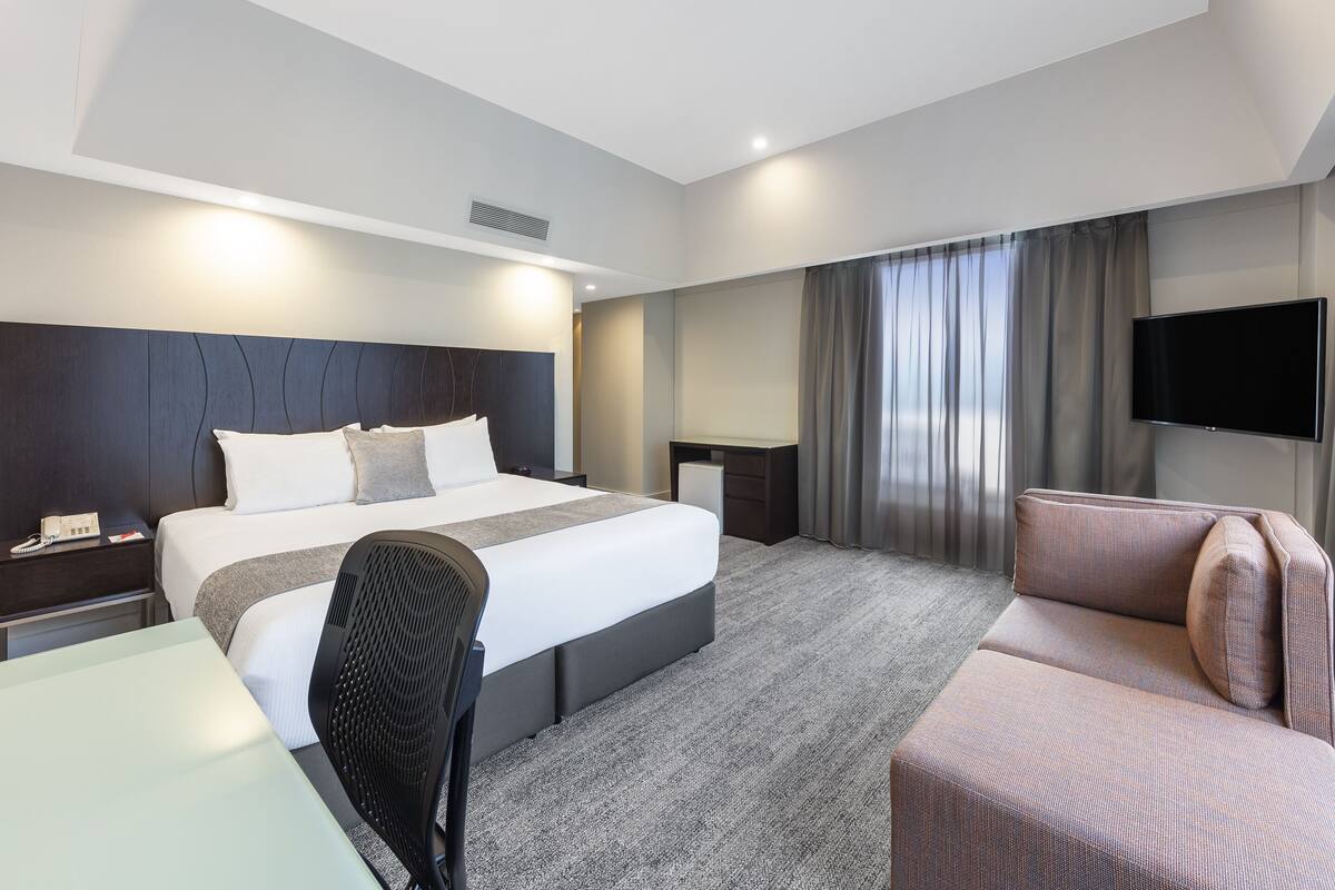Quarto de hotel moderno com uma cama grande, uma escrivaninha, uma cadeira, um sofá, televisão de tela plana e uma janela ampla com cortinas.