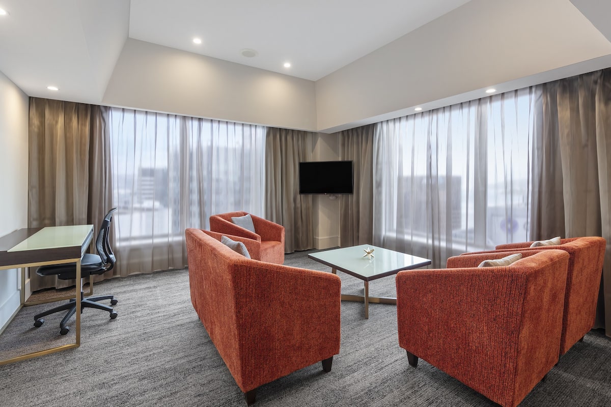 Quarto de hotel com uma janela grande, televisão de tela plana, escrivaninha e duas poltronas laranjas.