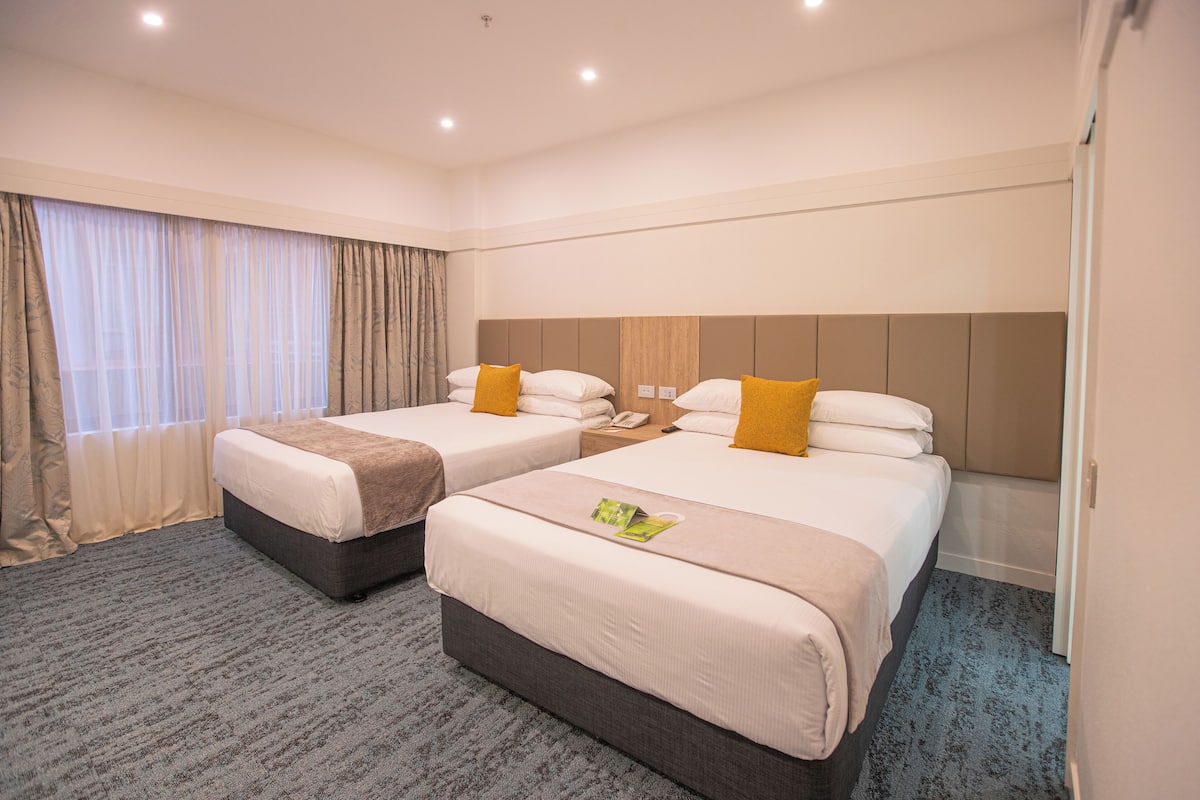 Quarto de hotel com duas camas, uma janela grande com cortinas, piso coberto de carpete e cabeceira de madeira.