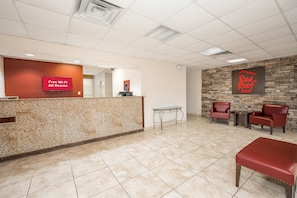 Uma área de recepção com um letreiro indicando 'Wi-Fi Gratuito em Todos os Quartos', uma parede de pedra com a sinalização 'Red Roof Inn' e um espaço de estar com duas cadeiras e um banco.