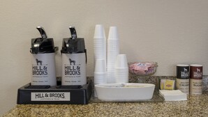 Estação de café com dois dosadores identificados como 'Hill & Brooks', uma bandeja com copos descartáveis, um recipiente com lenços de papel e recipientes com creme e açúcar.