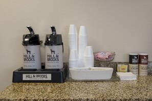 Estação de café com dois dosadores identificados como 'Hill & Brooks', uma bandeja com copos descartáveis, um recipiente com lenços de papel e recipientes com creme e açúcar.