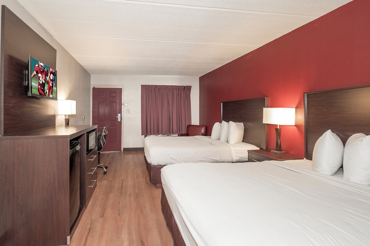 Quarto de hotel com duas camas, uma mesa de trabalho com televisão, parede vermelha e janela com cortinas.