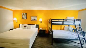 Quarto de hotel com duas camas, uma de casal e uma beliche, uma mesinha de cabeceira, um abajur e duas luminárias de parede.