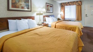 Quarto de hotel com duas camas, uma mesa de cabeceira com luminária, um quadro na parede e uma porta com aviso.