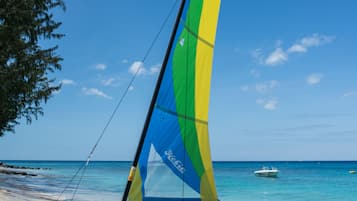 Um veleiro com vela azul e amarela numa praia de areia e águas azul-claras, com um barco pequeno ao longe.