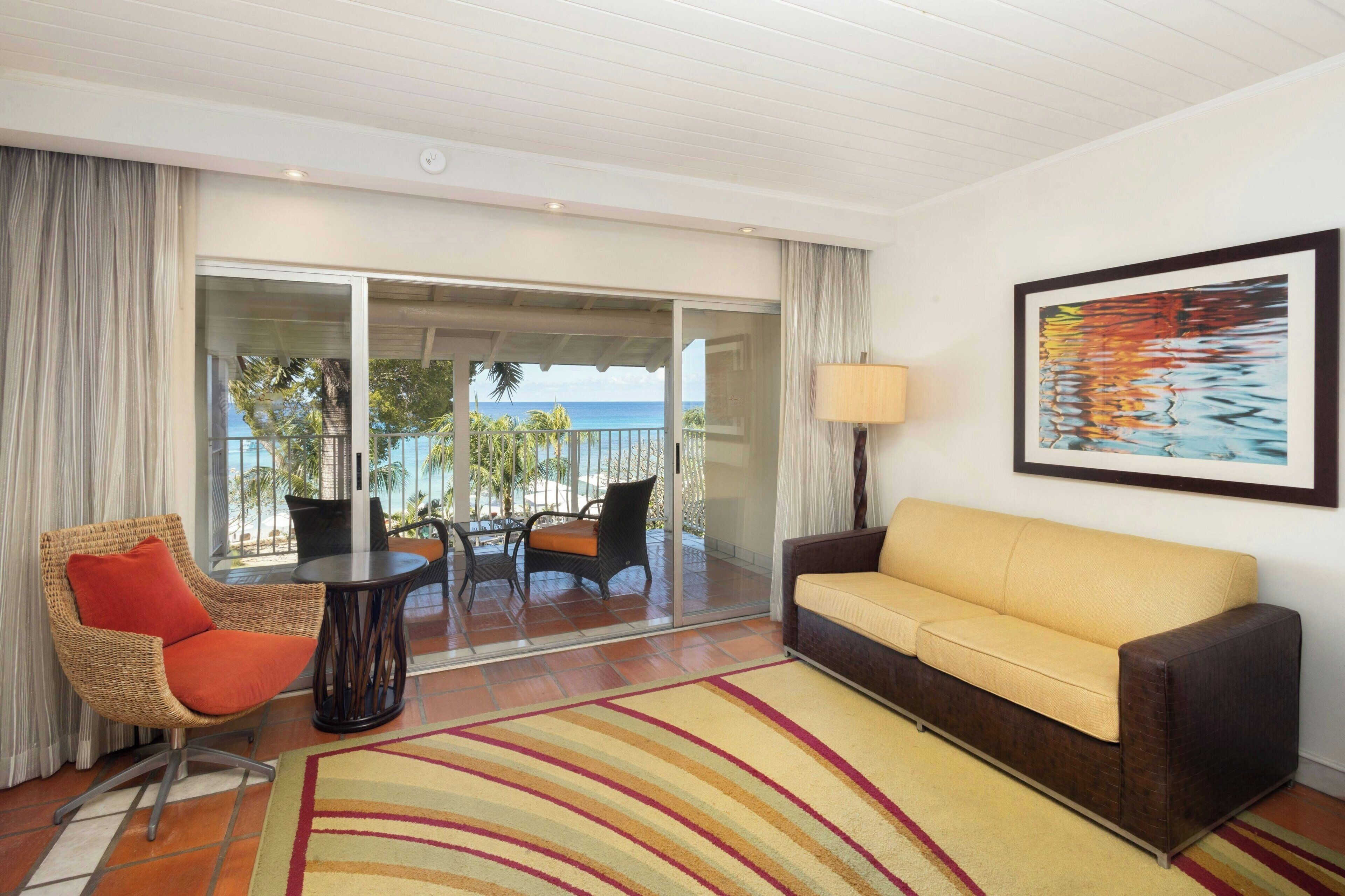 Quarto com sofá, cadeiras, mesa e vista para o oceano através de uma porta de vidro deslizante.