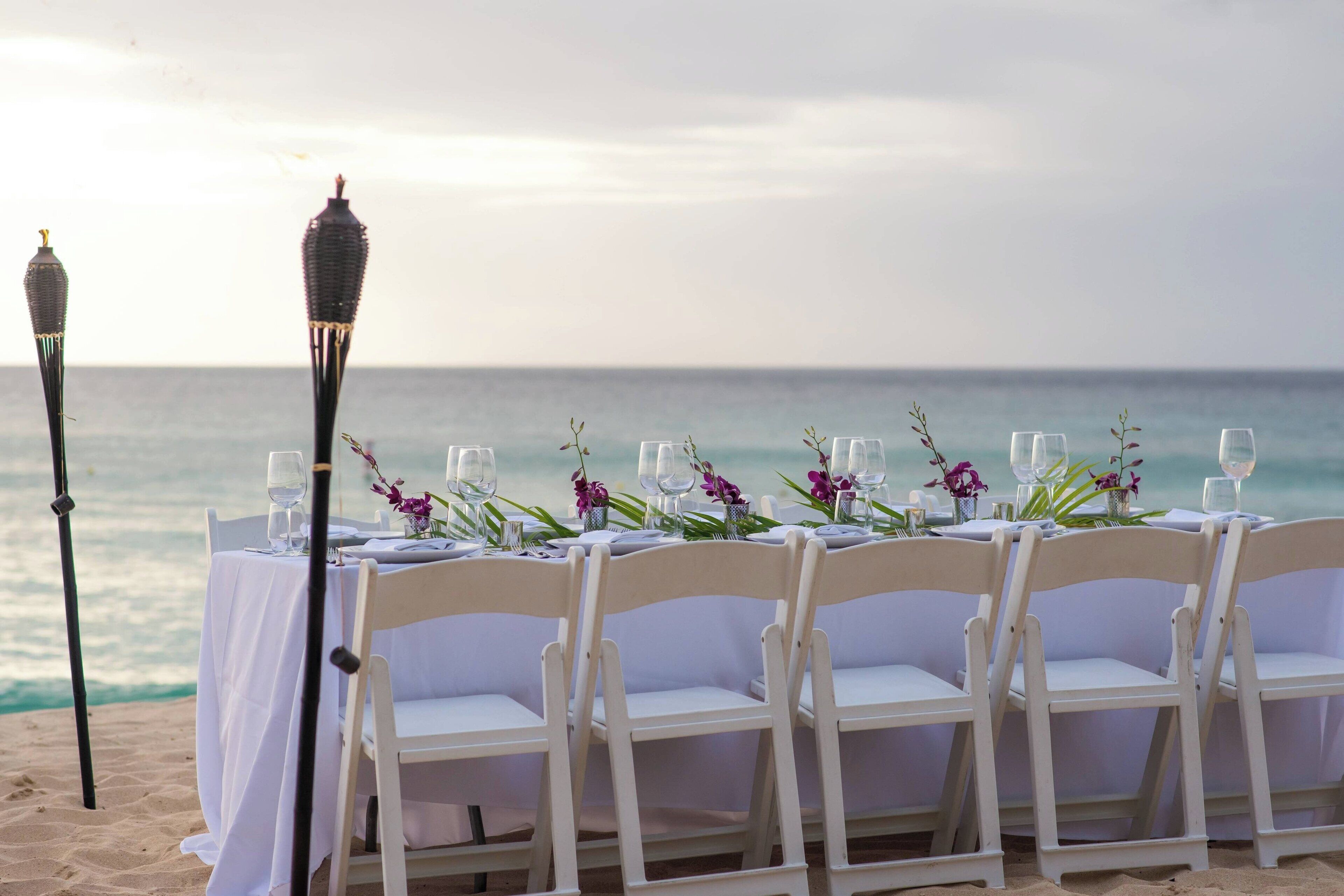 Disposição para refeições à beira-mar com cadeiras e mesas brancas, decorada com flores roxas e velas, com vista para o oceano.