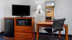 Quarto de hotel com TV de tela plana, micro-ondas, geladeira, escrivaninha de madeira, cadeira e luminária na parede.