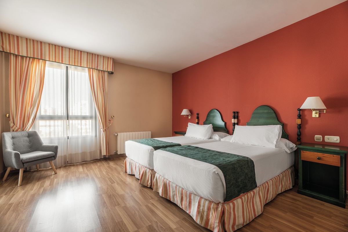 Quarto de hotel com uma cama, uma cadeira, uma mesinha de cabeceira e uma janela com cortinas.