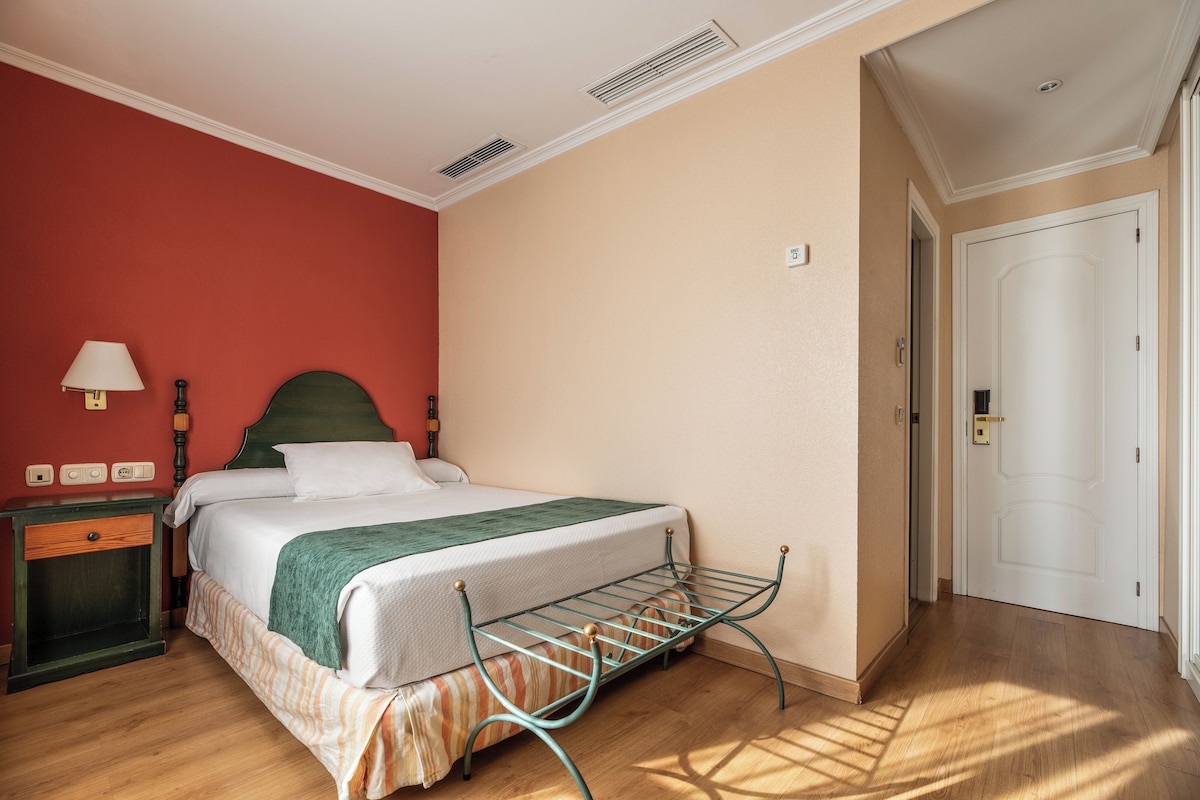 Quarto de hotel com cama, mesa de cabeceira, abajur, radiador e porta.
