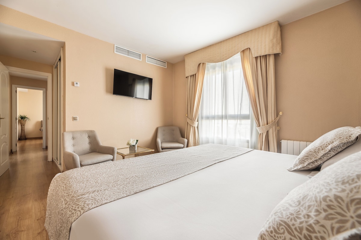 Quarto de hotel com uma cama grande, duas poltronas, uma televisão na parede e vista para o exterior pela janela.