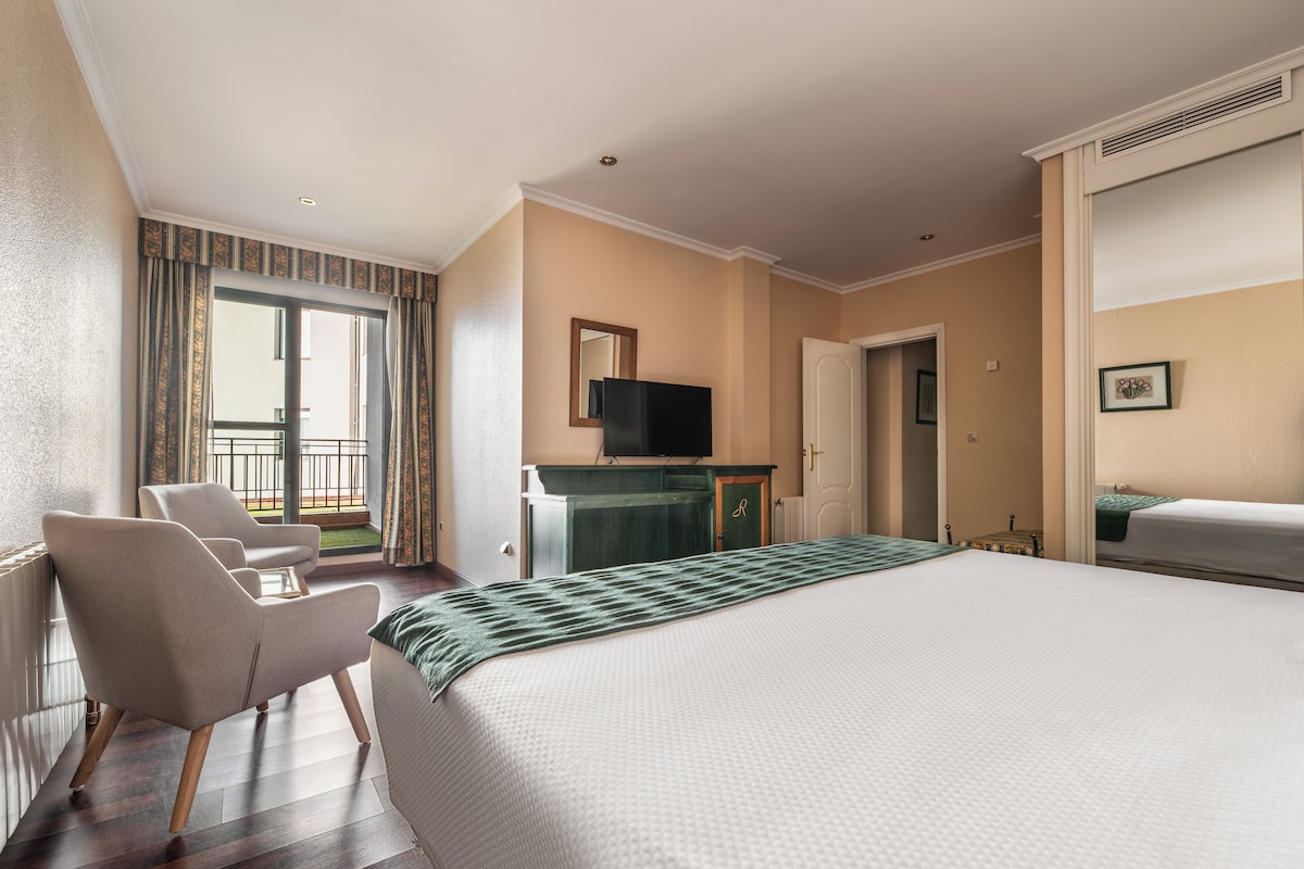 Quarto de hotel com uma cama grande, uma escrivaninha, uma cadeira, televisão e varanda com cortinas.