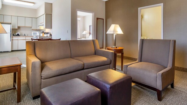 Quarto de hotel com sofá, poltrona, banquinho, uma pequena mesa, luminária e área de cozinha ao fundo.