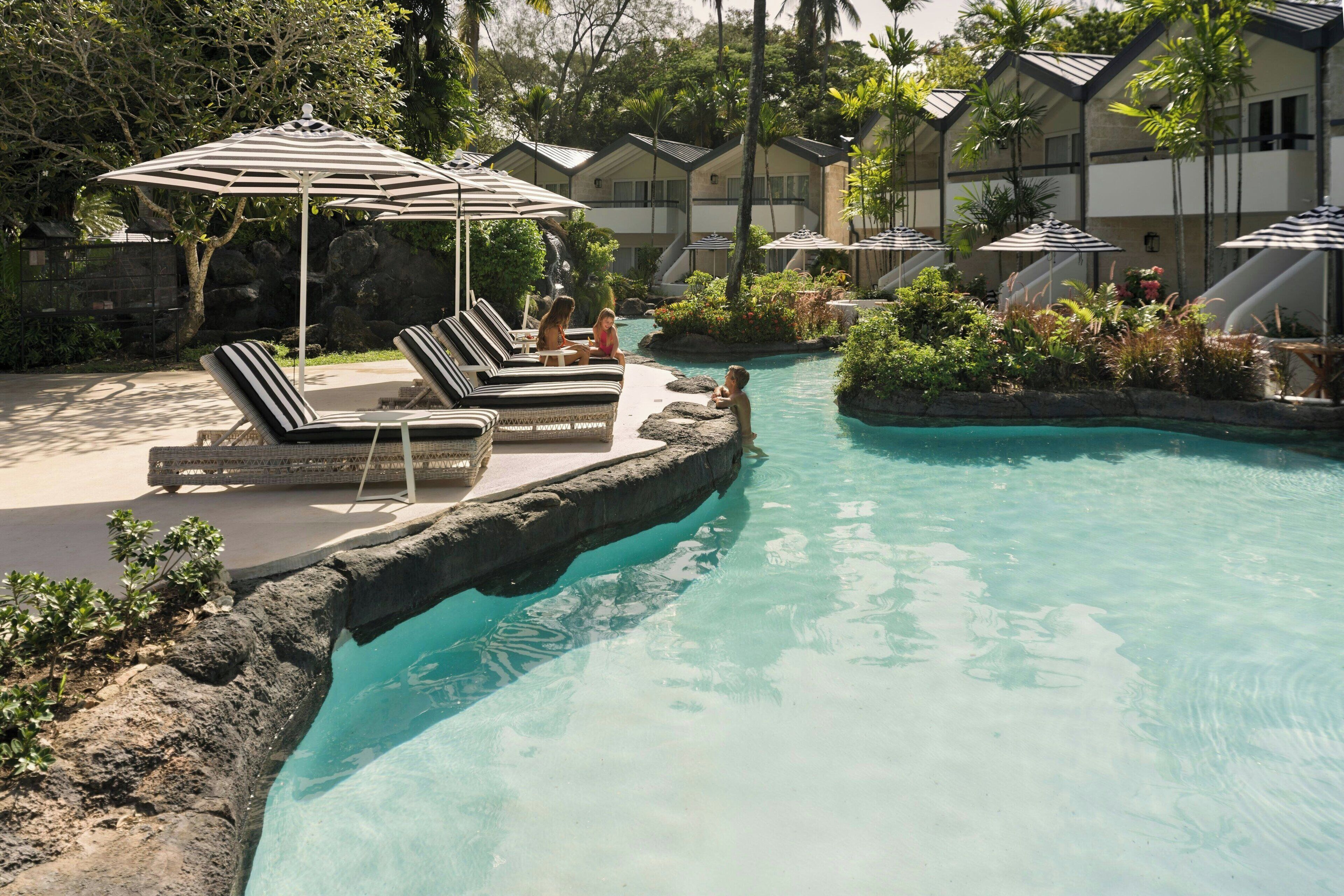 Uma área de piscina com espreguiçadeiras e guarda-sóis, cercada por vegetação e um edifício com várias varandas.