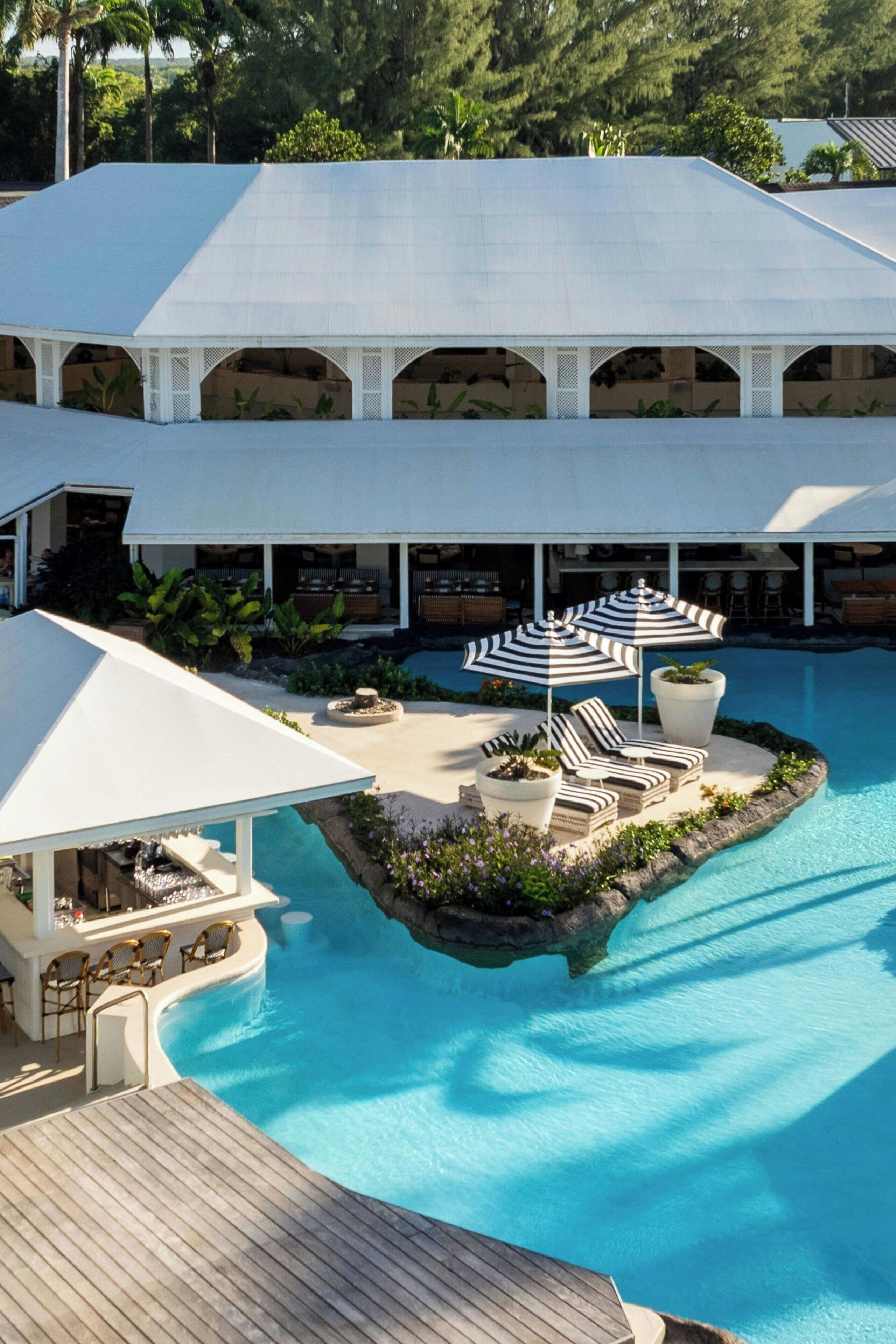 Uma área de piscina com espreguiçadeiras e guarda-sóis, rodeada por um alpendre coberto e um edifício com arcos.