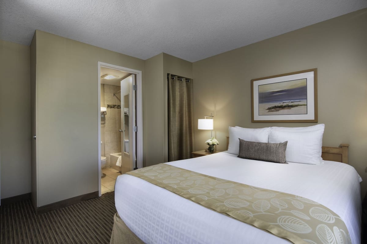 Quarto de hotel com cama, mesa de cabeceira com abajur, quadro na parede e porta aberta para um banheiro.