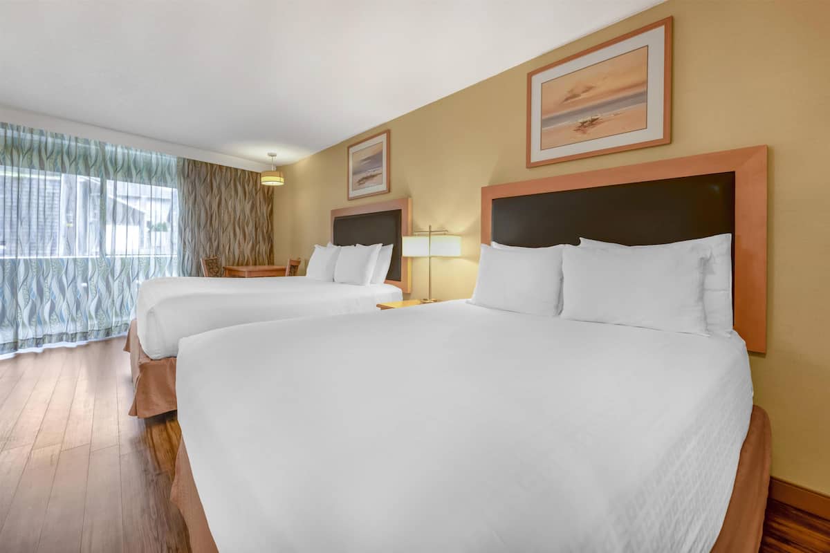Quarto de hotel com duas camas, uma janela com cortinas, uma luminária e duas fotos emolduradas na parede.