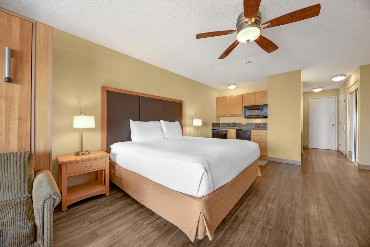 Quarto de hotel com cama, mesa de cabeceira, luminária, piso de madeira, ventilador de teto, cozinha compacta e uma porta.