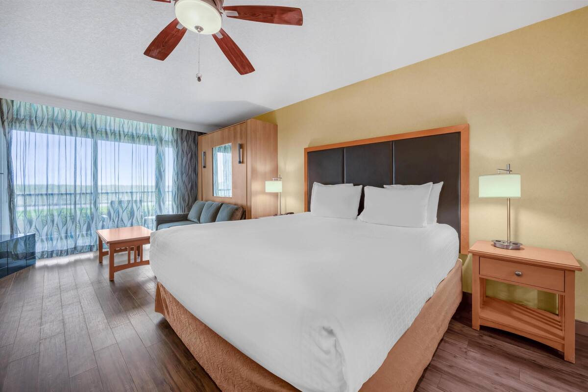 Quarto de hotel com uma cama grande, um ventilador de teto, uma mesa, uma cadeira, uma mesinha de cabeceira e vista para o exterior.