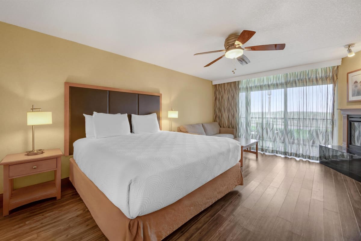 Quarto de hotel com uma cama grande, duas mesas de cabeceira com luminárias, um ventilador de teto, vista para o exterior através de uma janela com cortinas e uma lareira.