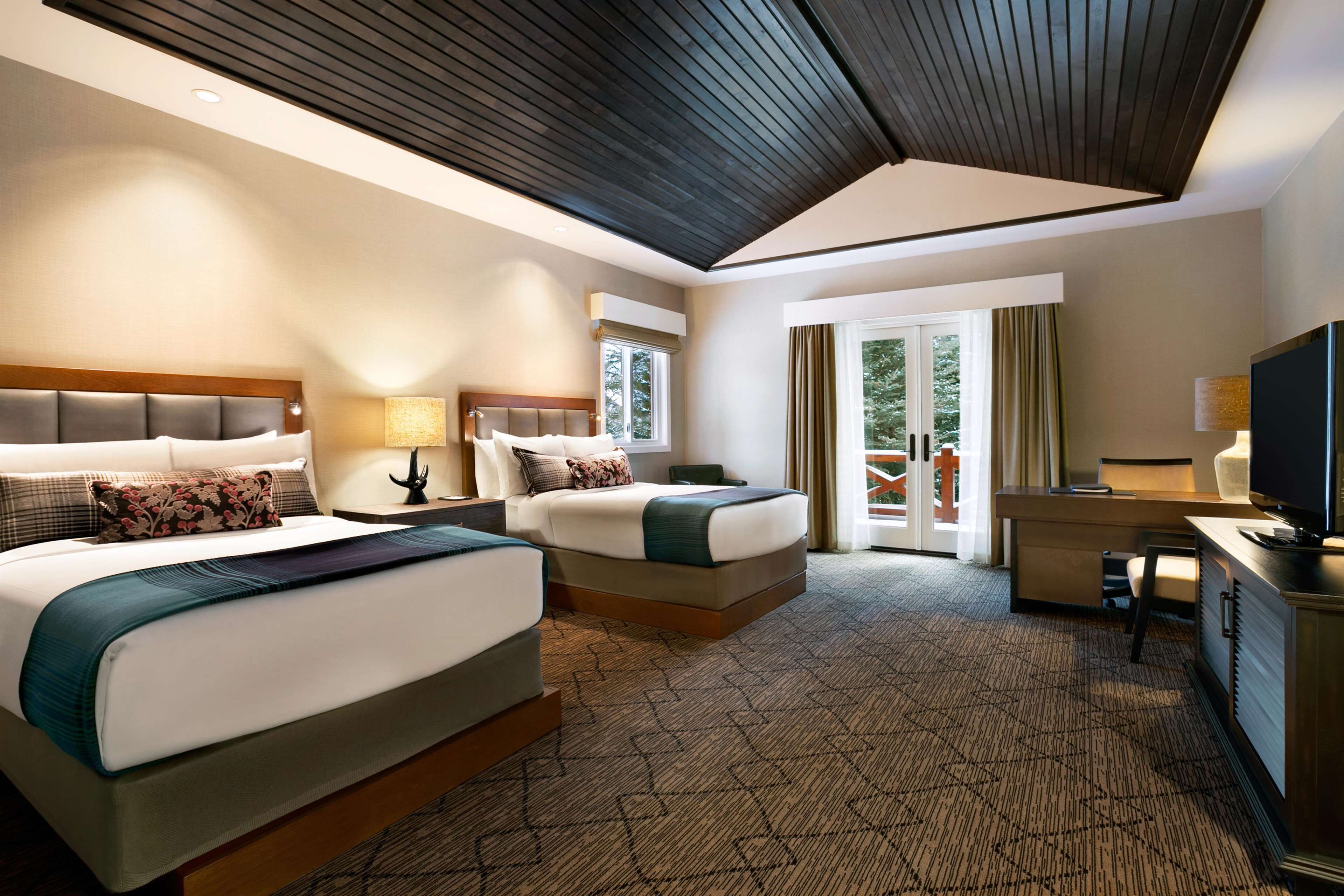 Quarto de hotel com duas camas, uma escrivaninha, televisão e varanda com vista para árvores.