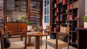 Um quarto com uma mesa redonda de madeira, rodeada por cadeiras, uma estante com livros e objetos decorativos, e um suporte para vinhos com garrafas.