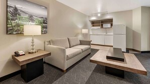 Quarto de hotel moderno com sofá, mesa de centro, escrivaninha com luminária, pintura na parede e cozinha compacta ao fundo.